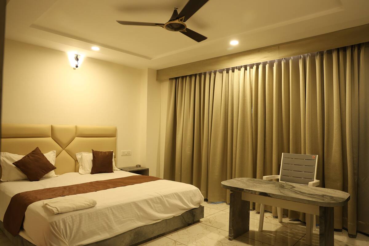 Quarto de hotel com cama, escrivaninha, cadeira, ventilador de teto e cortinas.