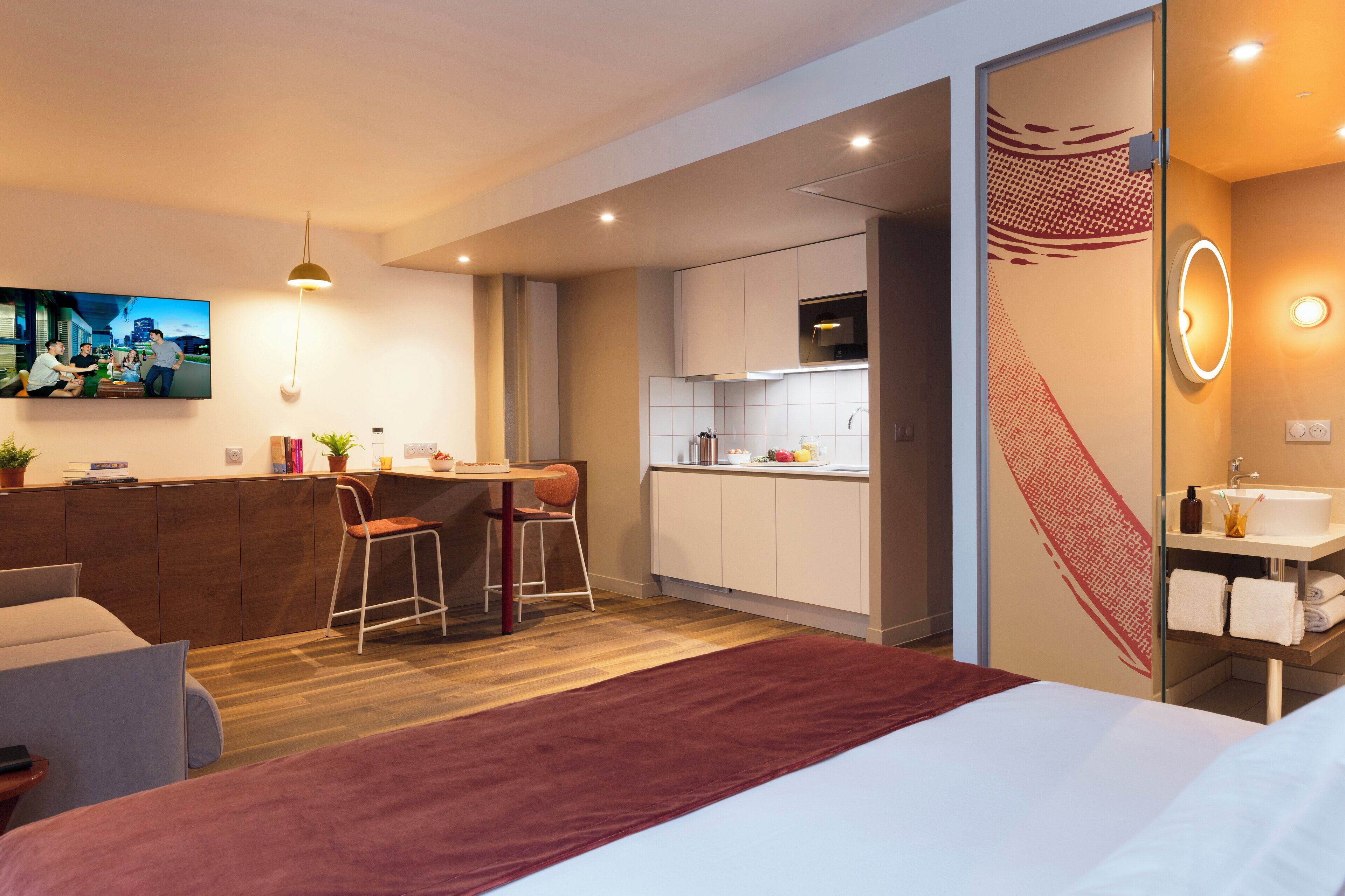 Quarto de hotel moderno com cama, escrivaninha, cadeira, pequena cozinha e banheiro com espelho e pia.