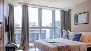 Quarto de hotel com uma grande janela, cama com roupa de cama branca e vista para a cidade.