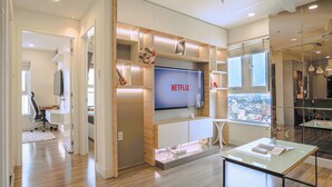 Uma sala de estar moderna com uma grande TV de tela plana exibindo 'NETFLIX', um rack de madeira, uma mesa de centro de vidro e vista para a cidade através de grandes janelas.
