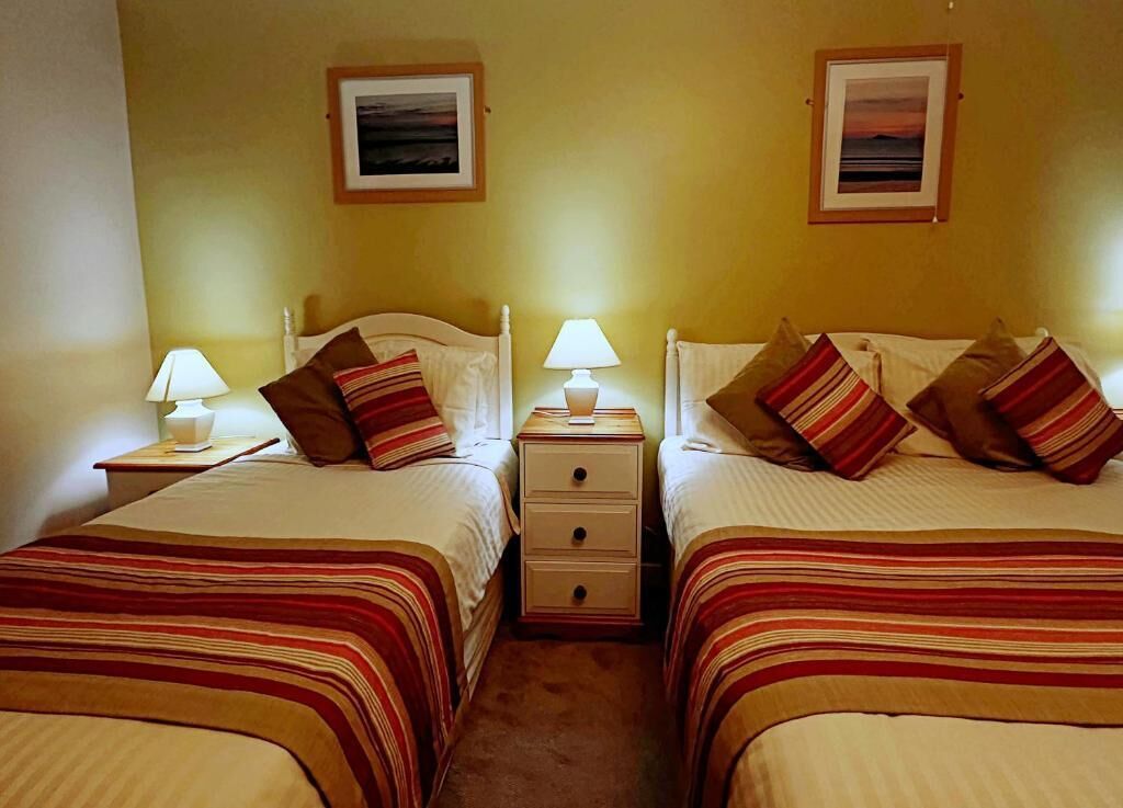 Quarto de hotel com duas camas, cada uma com roupa de cama e almofadas listradas, uma mesa de cabeceira com abajur e uma pequena gaveta.