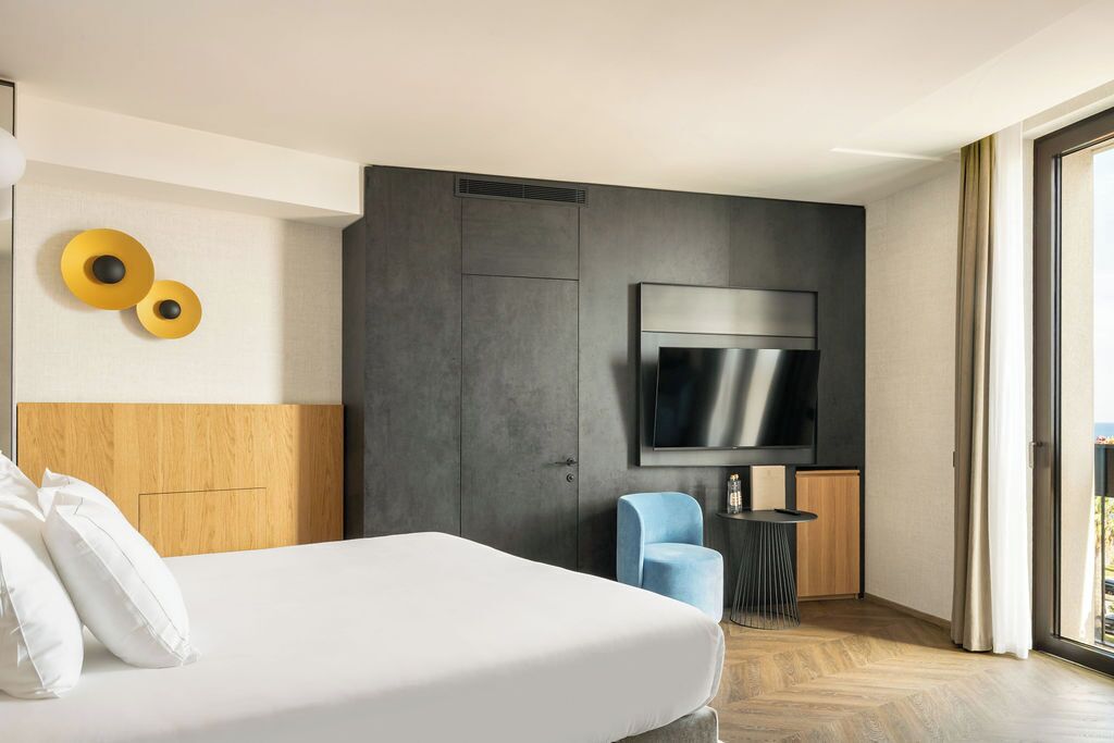 Quarto de hotel moderno com uma cama grande, uma televisão de tela plana na parede, uma mesinha redonda e uma cadeira.
