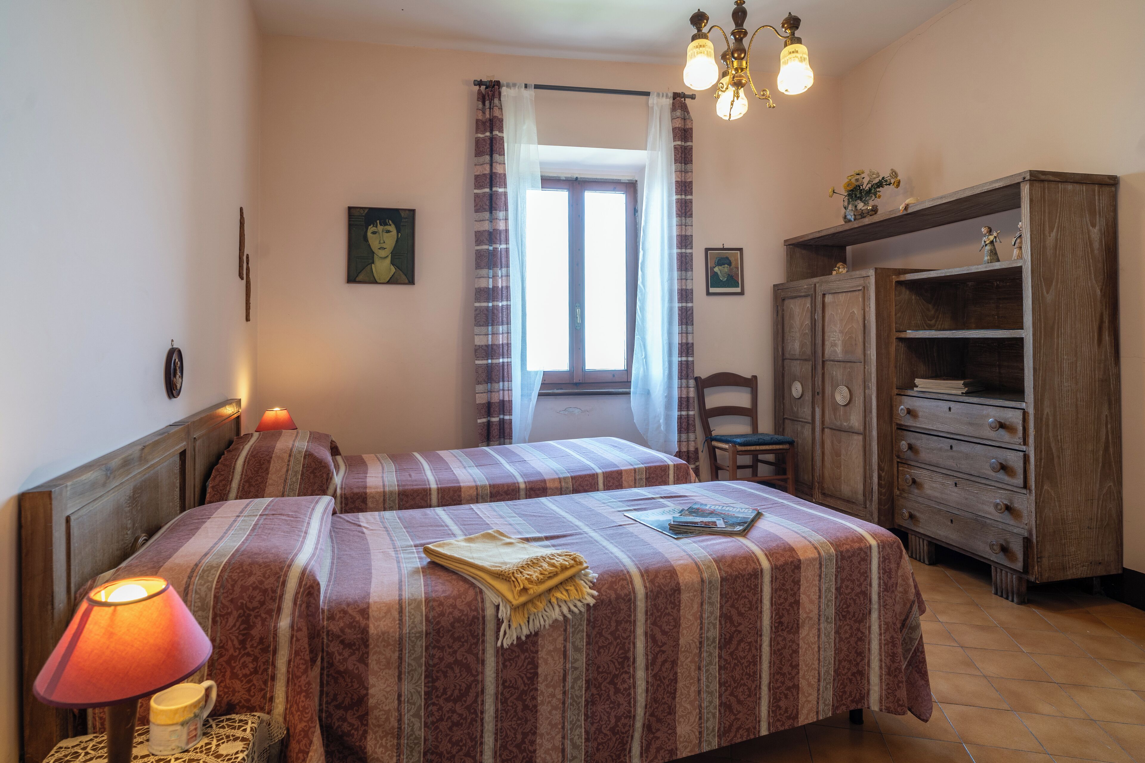 Quarto com duas camas, guarda-roupa de madeira, janela com cortinas e mesa de cabeceira com abajur.