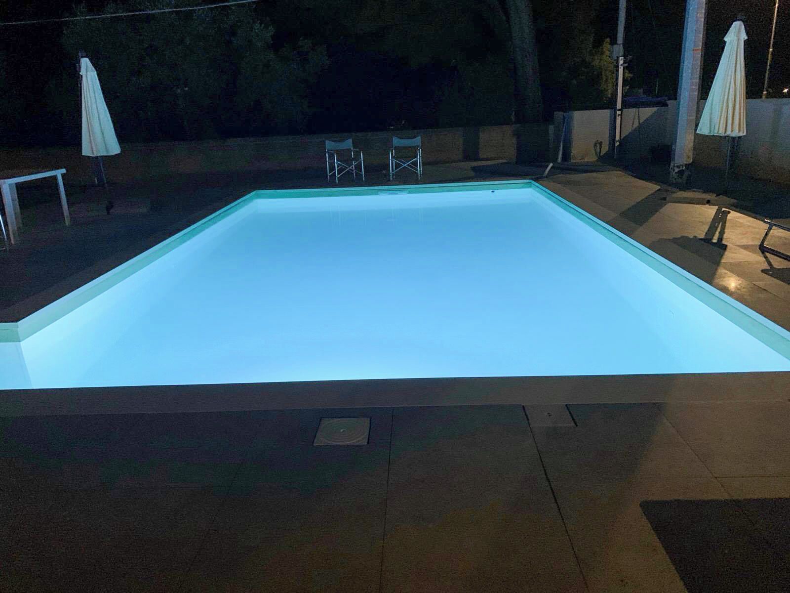 Piscina retangular com borda iluminada, cercada por um deck ladrilhado e duas ombrelhas brancas.