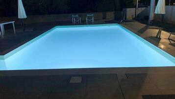 Piscina retangular com borda iluminada, cercada por um deck ladrilhado e duas ombrelhas brancas.