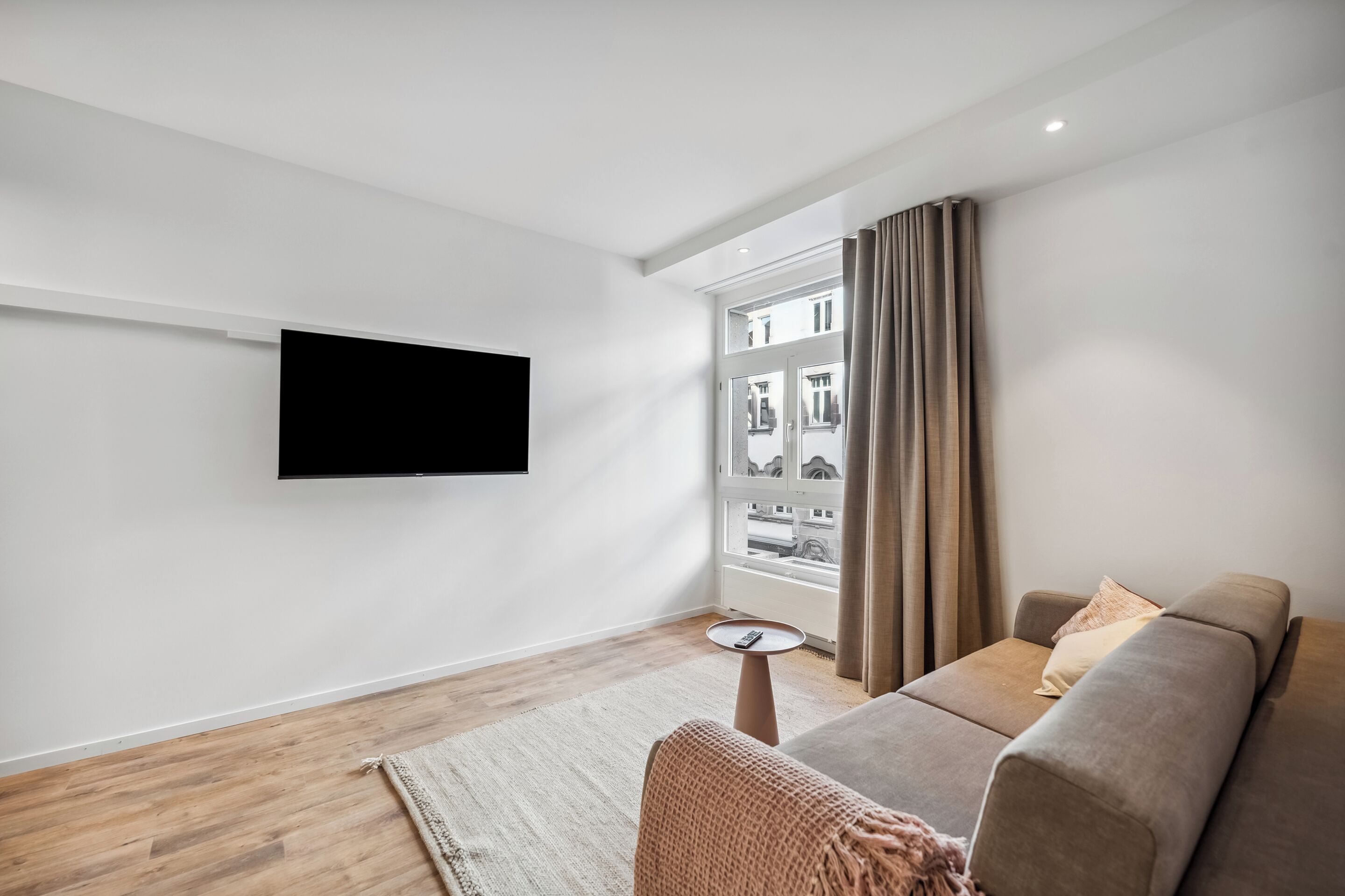 Uma sala de estar moderna com uma TV de tela plana na parede, um sofá com almofadas, uma mesinha redonda com um controle remoto e vista para edifícios através de grandes janelas.