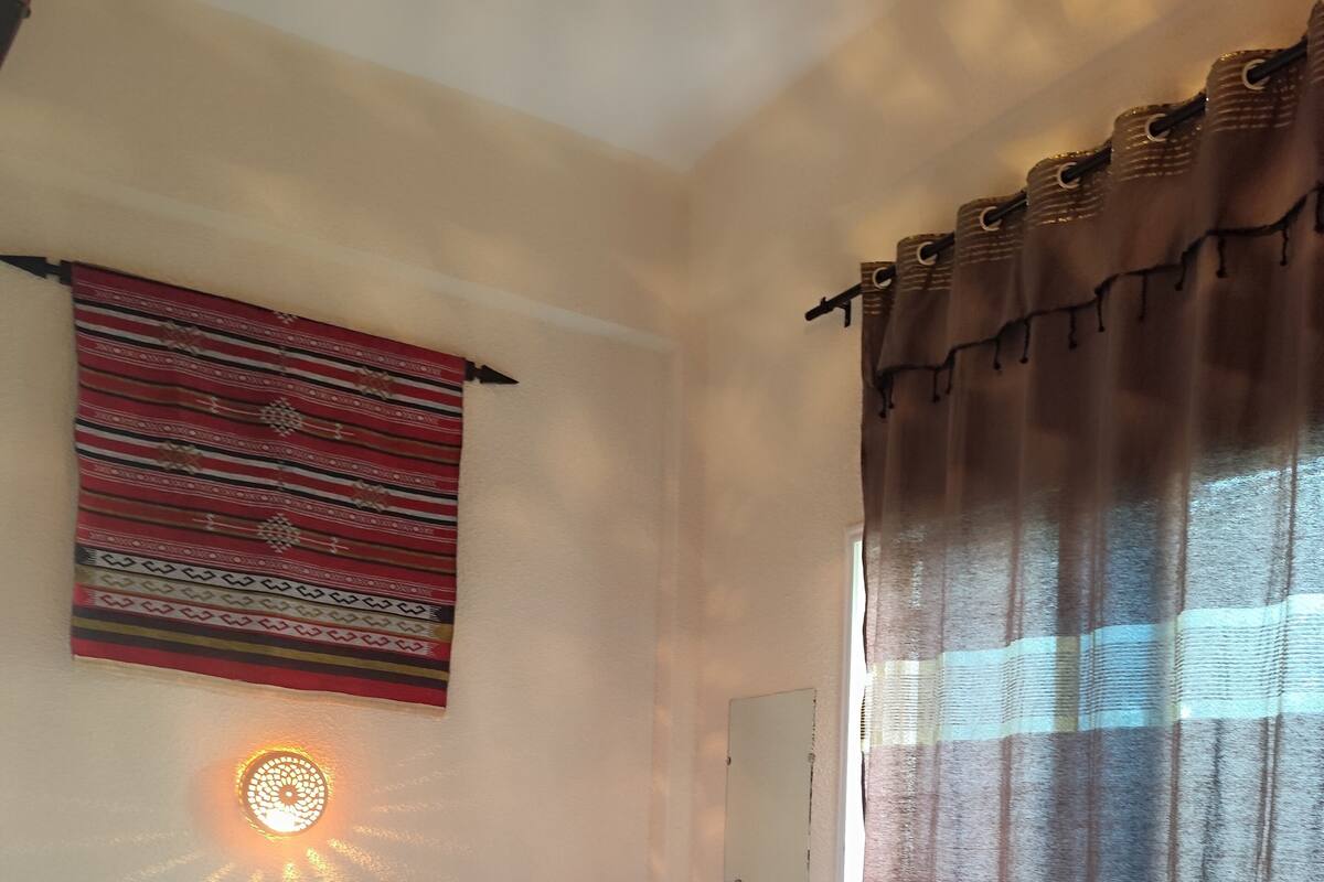 Quarto com cama, mesa de cabeceira, luminária na parede, pia e janela com cortinas.