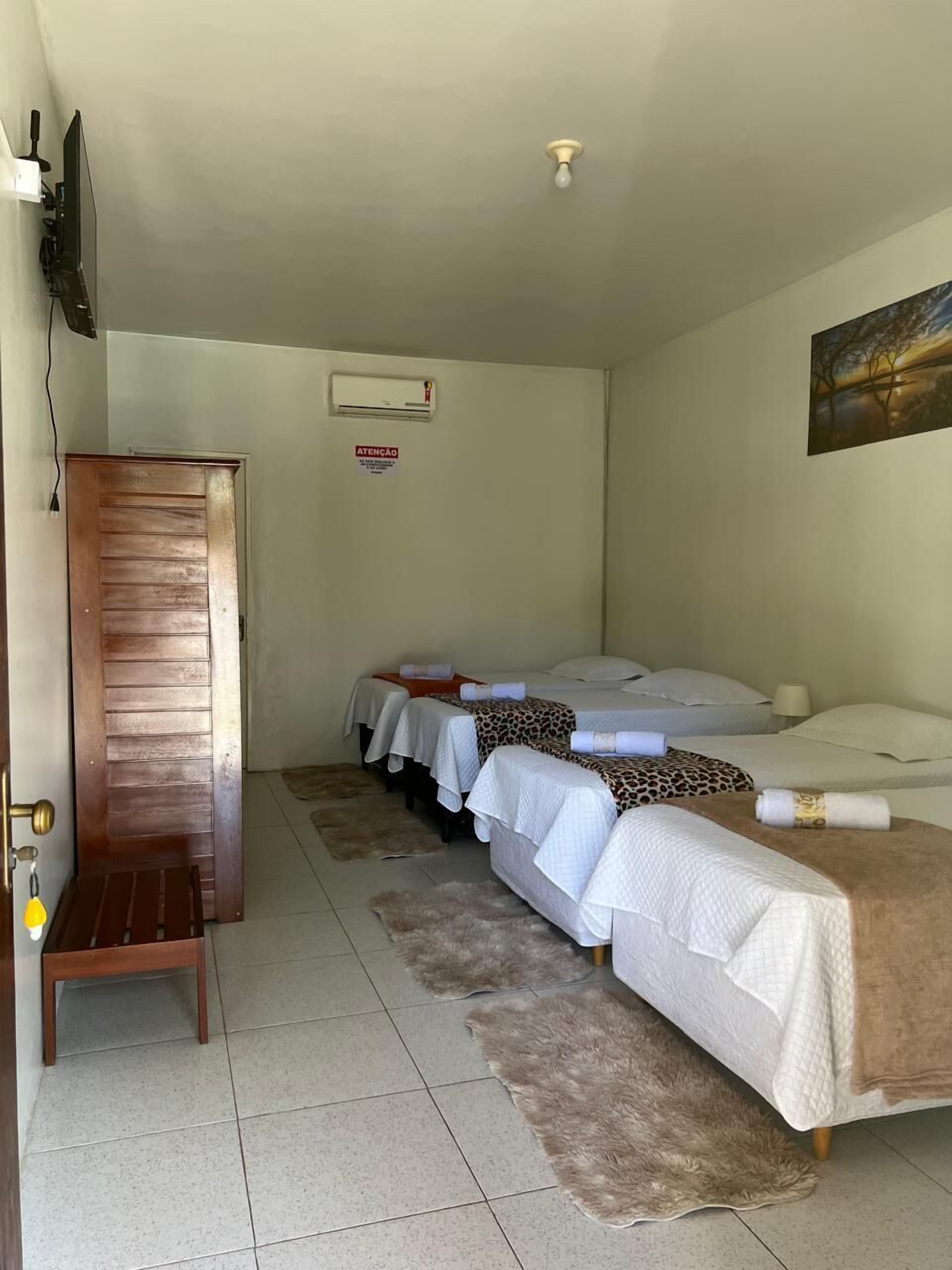 Quarto de hotel com três camas, porta de madeira, ventilador de teto, ar condicionado e um quadro na parede.