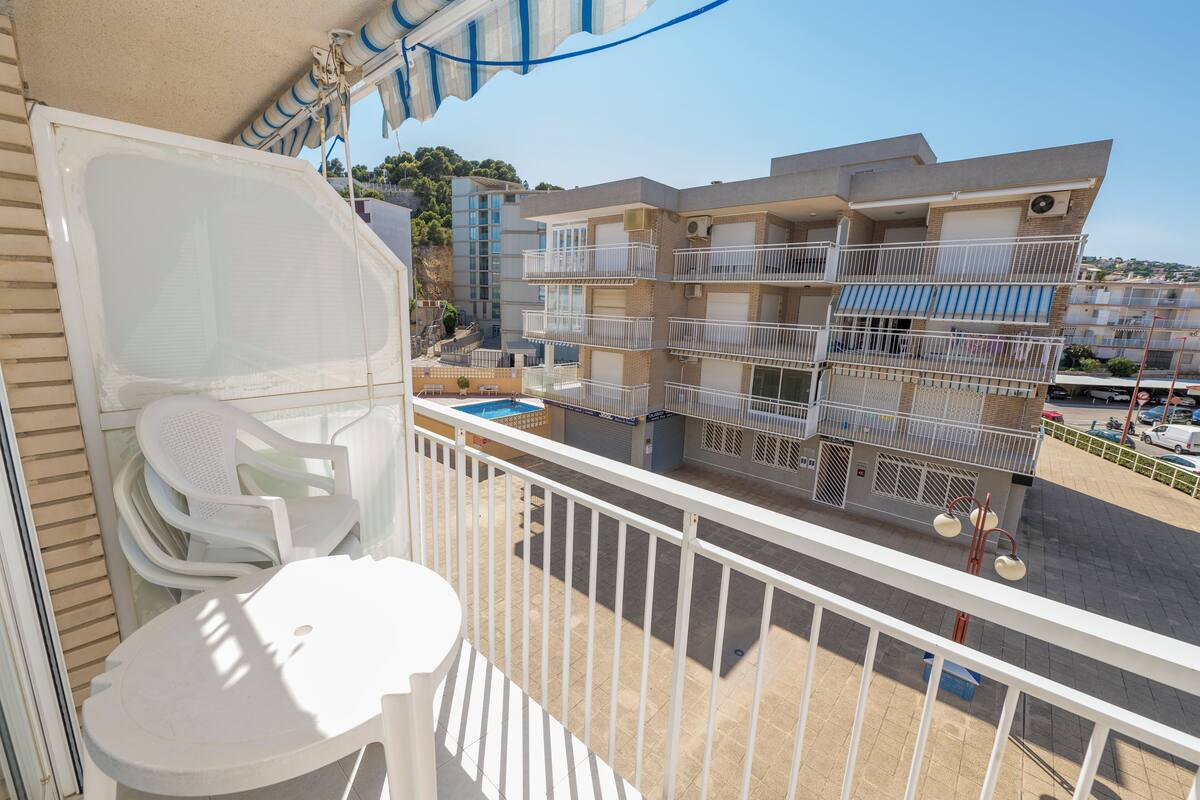 Uma varanda com mesa e cadeiras brancas, piscina e vista para edifícios altos.