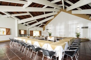 Uma sala de conferências com mesas compridas e cadeiras, paredes brancas e teto alto com vigas aparentes.