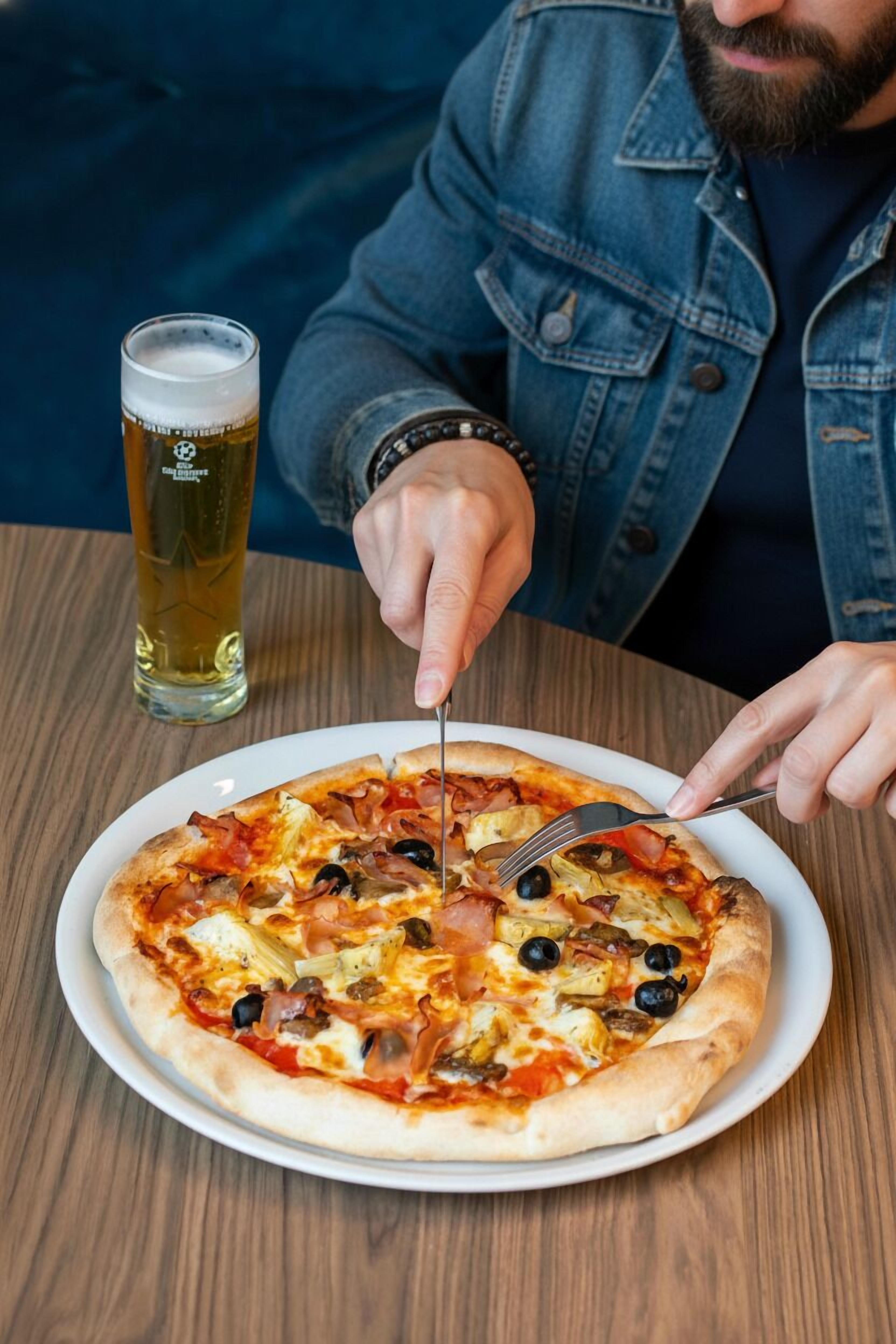 Uma pessoa cortando uma pizza com garfo e faca sobre uma mesa de madeira.