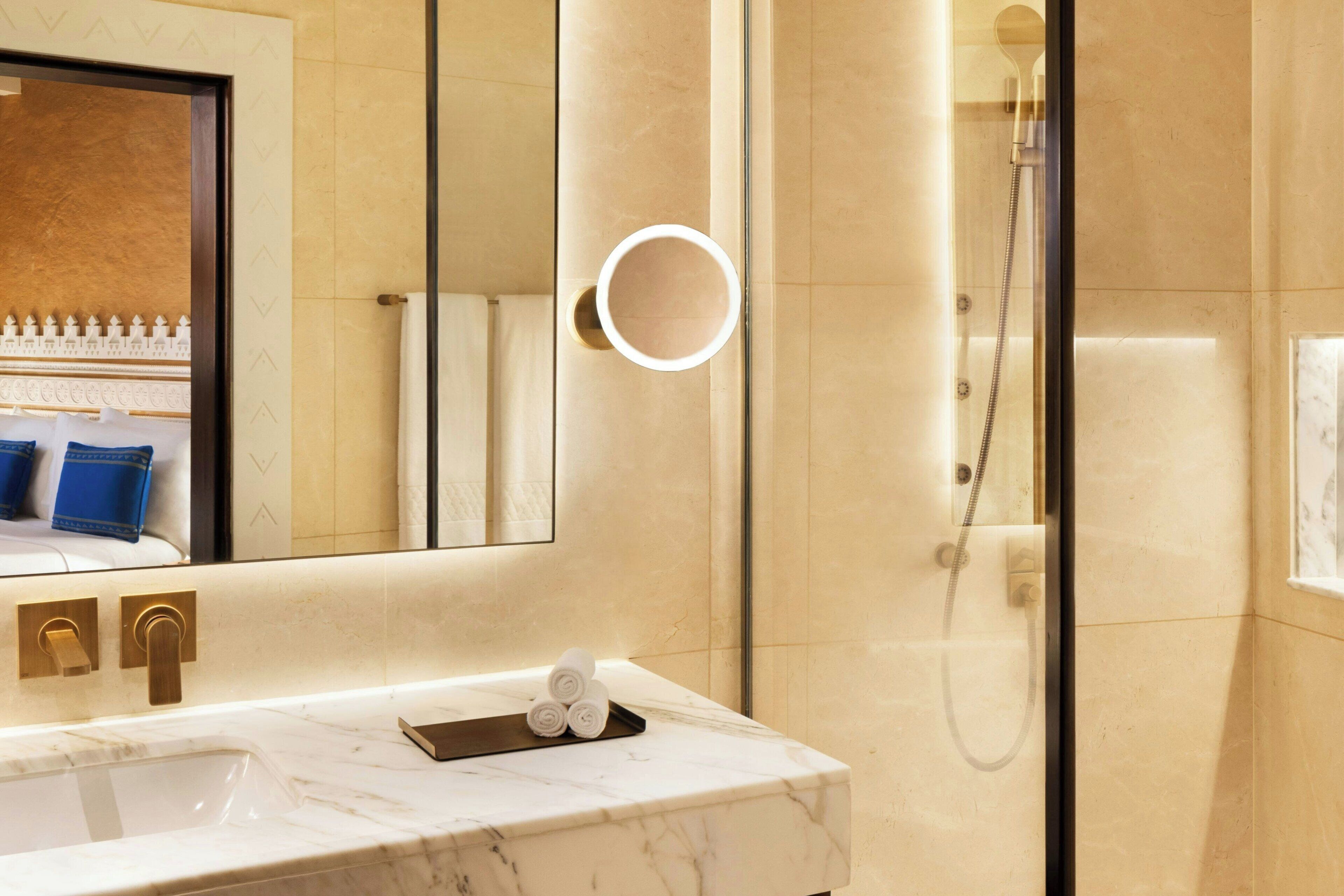 Um banheiro moderno com pia de mármore, chuveiro com box de vidro e um espelho grande com iluminação circular.