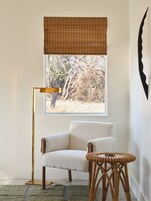 Um quarto com uma janela grande, uma poltrona branca, um abajur e um banquinho de fibra.
