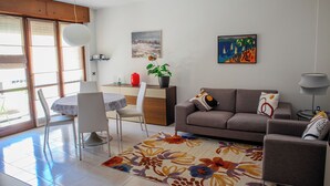 Uma sala de estar moderna com um sofá, uma mesa de jantar com cadeiras, uma mesa de centro e uma janela grande com persianas.