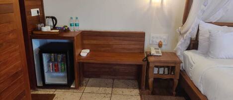 Quarto de hotel compacto com um banco de madeira, um pequeno frigorífico, uma mesa de cabeceira com telefone e uma cama com dossel.