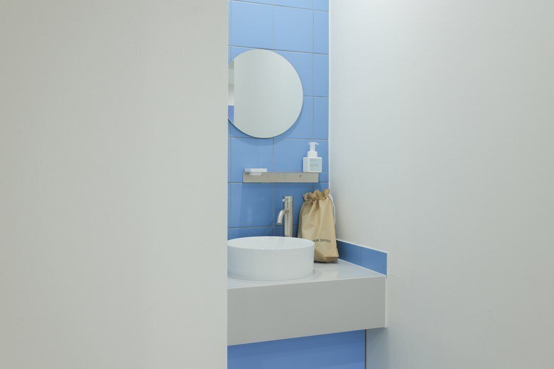 Casa de banho com lavatório branco, espelho redondo, porta-toalhas e pequena prateleira com produtos de higiene pessoal.