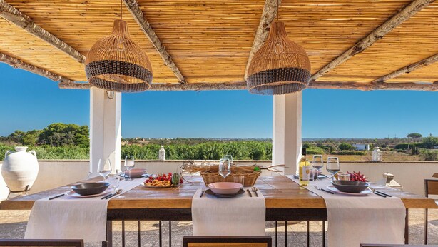 Uma área de refeições ao ar livre com uma mesa posta para dois, sob um telhado de palha, com vista para uma paisagem deslumbrante.
