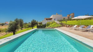 Uma piscina retangular com água azul cristalina, rodeada por um deck pavimentado. Ao fundo, vê-se uma casa moderna com fachada de pedra e um arco de pedra.