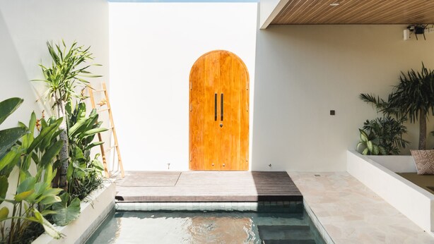 Piscina com porta de madeira ao fundo, rodeada por paredes brancas e plantas em vasos.
