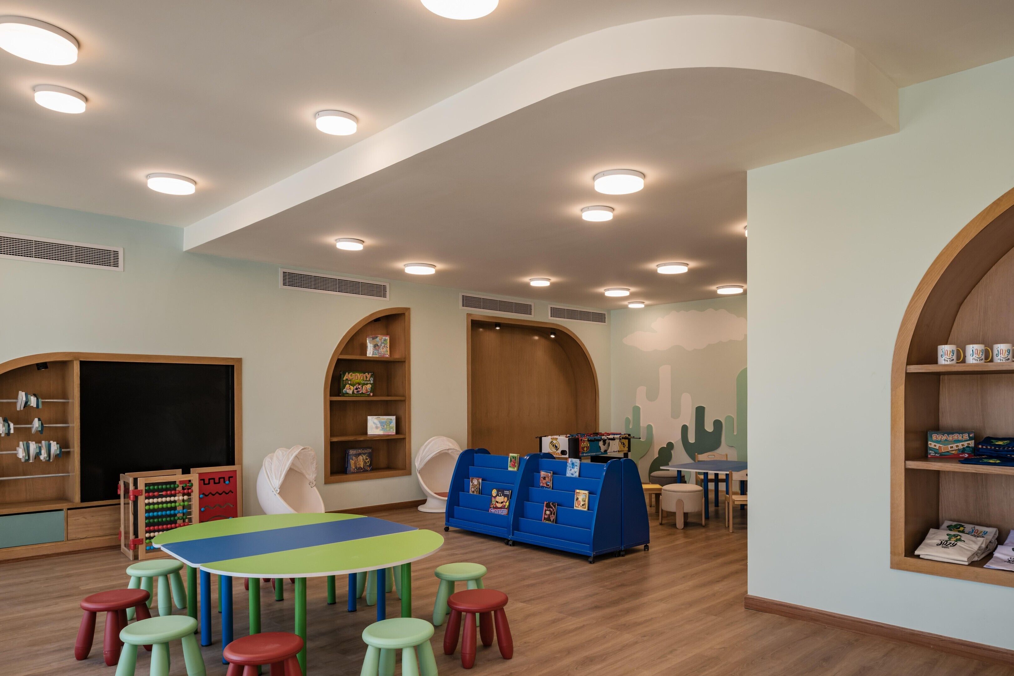 Uma sala de brincar infantil com uma mesa redonda, banquetas, prateleiras com brinquedos e um mural na parede.