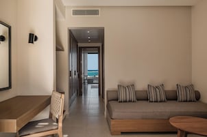 Quarto de hotel moderno com uma escrivaninha de madeira, uma cadeira estofada, um sofá com almofadas listradas e vista para o oceano através de uma porta aberta.