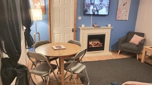 Uma sala de estar aconchegante com lareira, uma mesa de jantar redonda com cadeiras, uma televisão na parede e uma poltrona confortável.