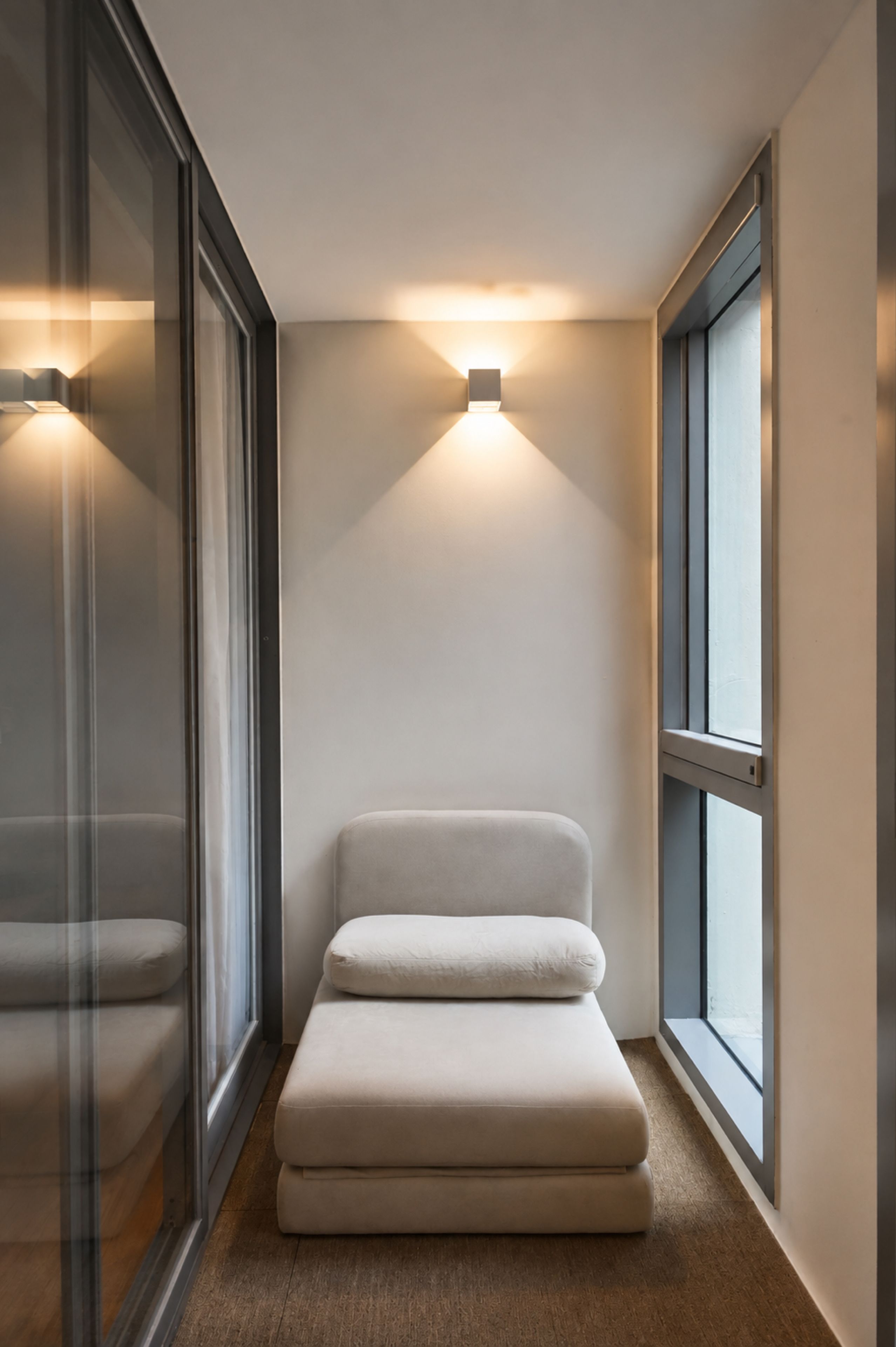 Quarto estreito com um banco, uma janela e duas luzes fixadas na parede.