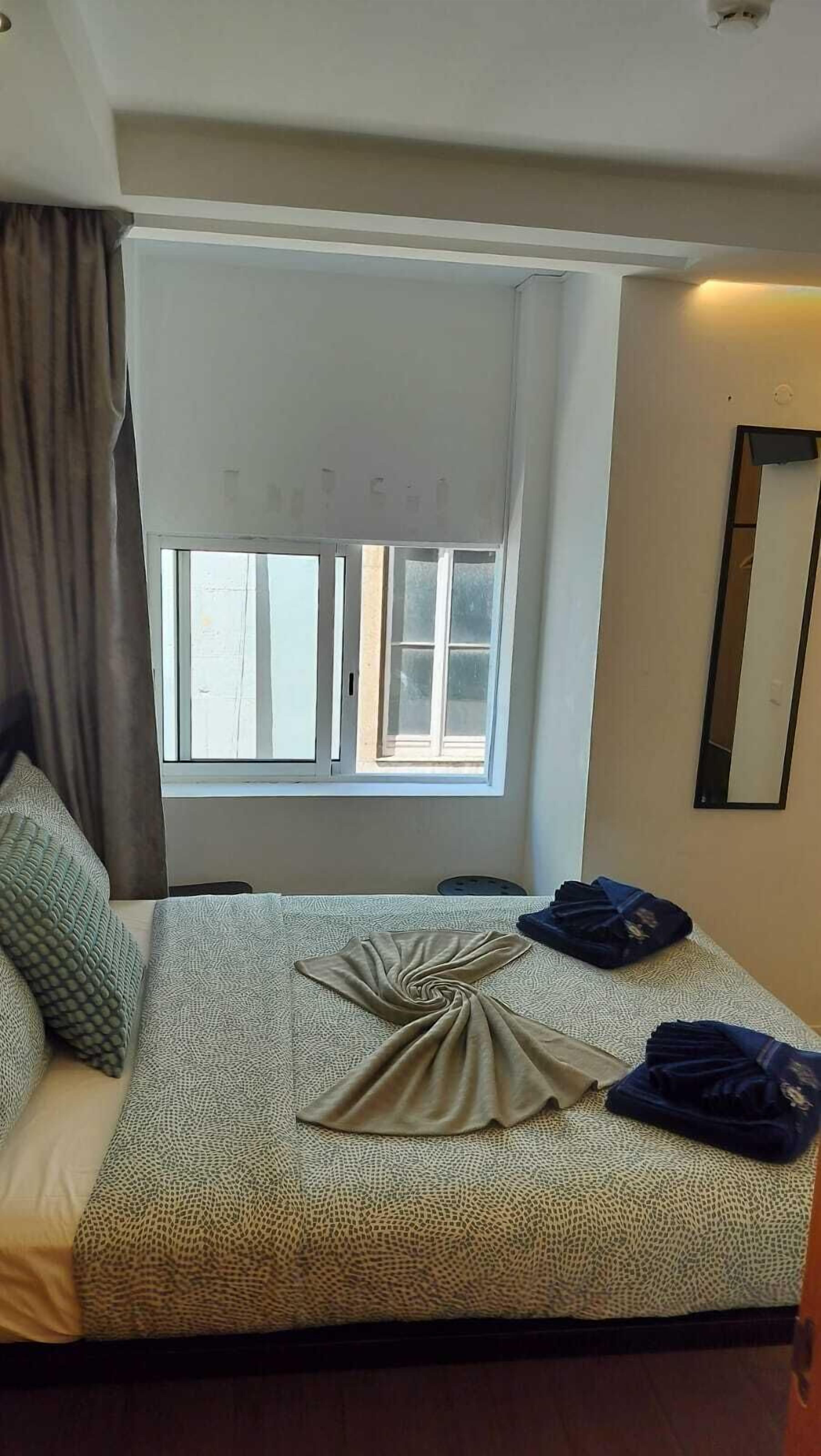 Quarto de hotel com cama, janela com persianas, espelho na parede e uma pequena mesa.