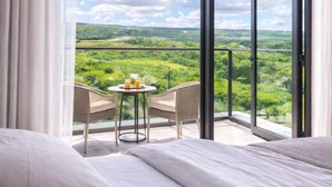 Quarto de hotel com uma grande porta de vidro deslizante que dá para uma varanda com mesa e cadeiras, proporcionando vista de uma paisagem verde.