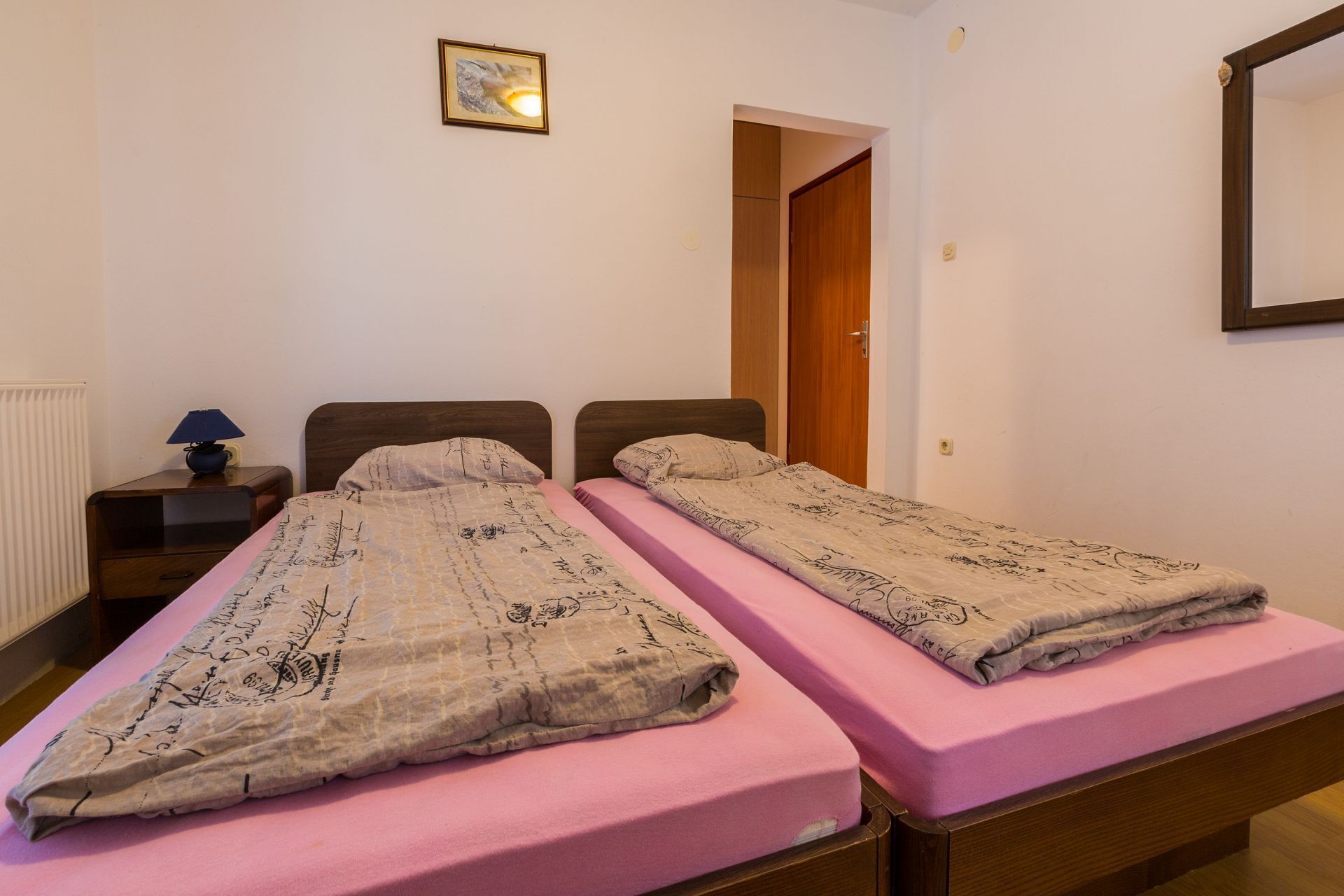 Quarto com duas camas de solteiro, mesa de cabeceira com abajur, quadro pendurado na parede e porta que dá para outro cômodo.