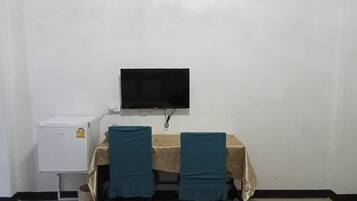 Um quarto pequeno com uma televisão fixada na parede, uma geladeira branca e duas cadeiras com capas azuis.