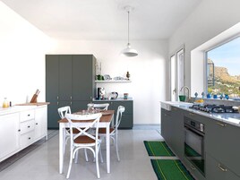 Cozinha moderna com armários brancos, mesa de jantar com cadeiras, luminária pendente e vista para o exterior pela janela.