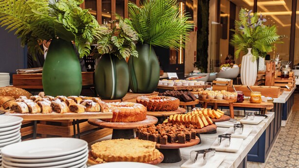 Buffet com diversos doces e pastelaria, grandes vasos verdes e uma longa mesa de serviço.