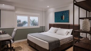 Quarto moderno com uma cama grande, uma escrivaninha e uma cadeira. Tem uma janela com vista para palmeiras.