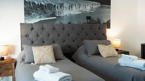 Uma cama de casal com roupa de cama cinza e cabeceira estofada, duas luminárias de apoio e um quadro grande emoldurado de uma geleira.