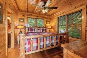 Quarto de cabana de madeira com cama, duas janelas, ventilador de teto e mesa de cabeceira.