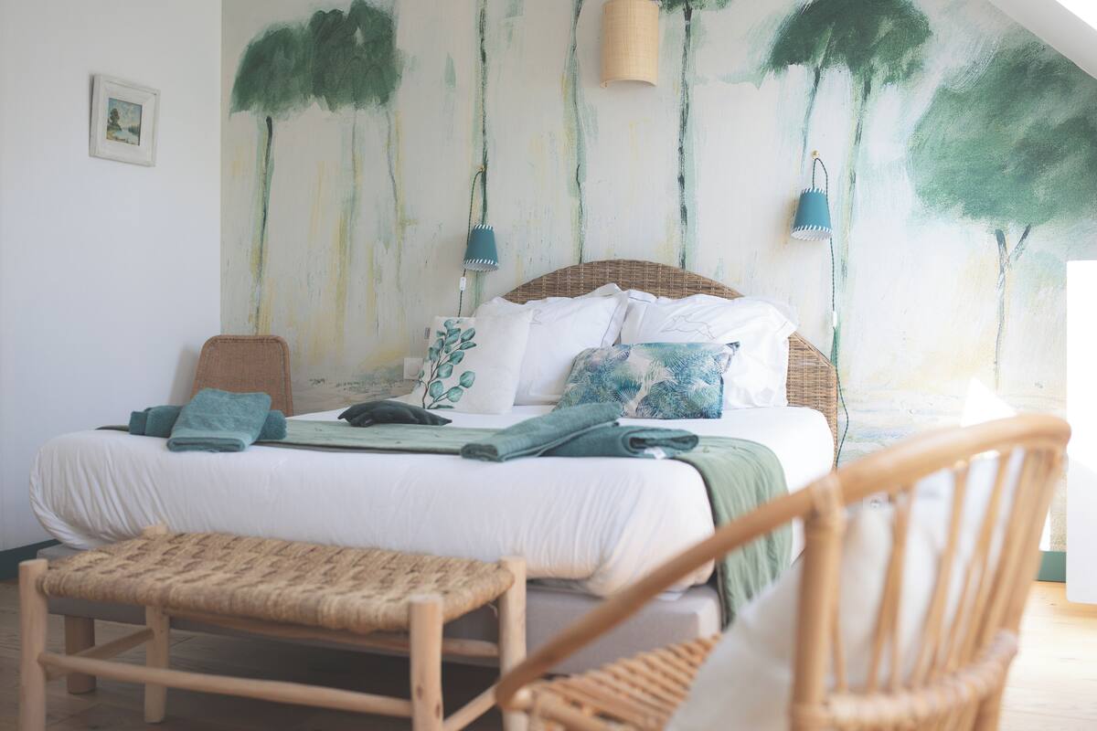 Quarto com cama, uma cadeira e parede com pintura de árvore.
