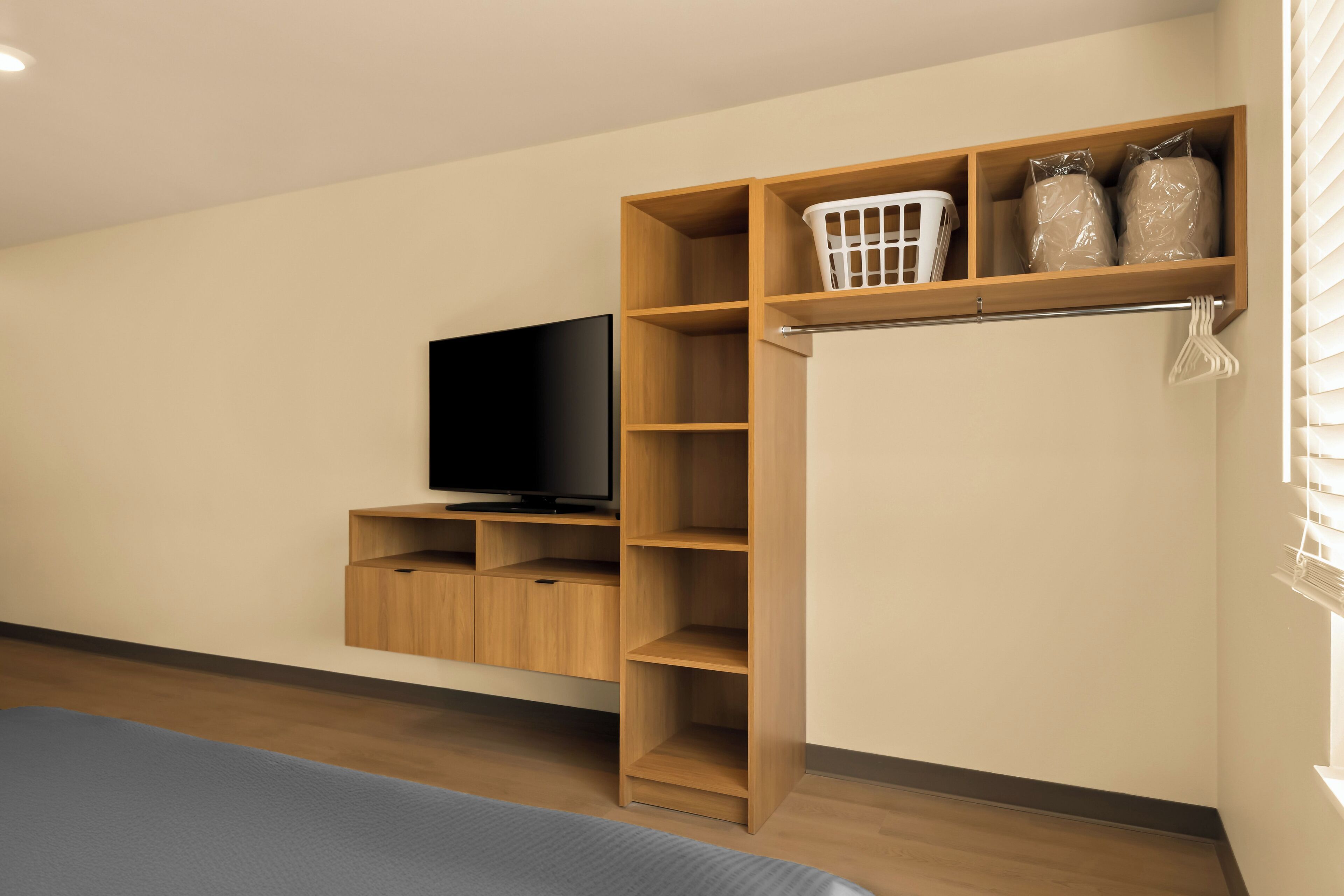 Quarto moderno com televisão de tela plana fixada em prateleira de madeira, cama com colchão azul e guarda-roupa de madeira com espaço para pendurar roupas.