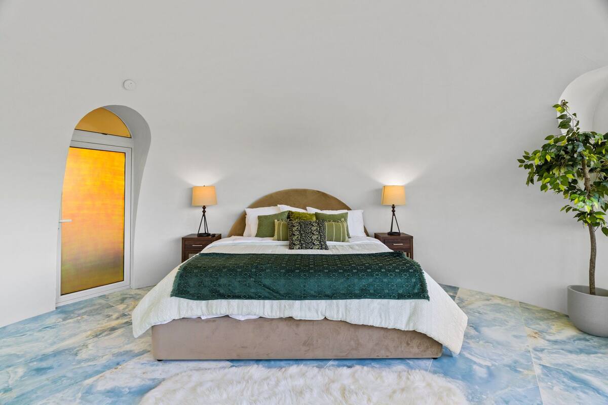 Quarto com cama, mesas de cabeceira, planta em vaso e porta com painel de vidro jateado.