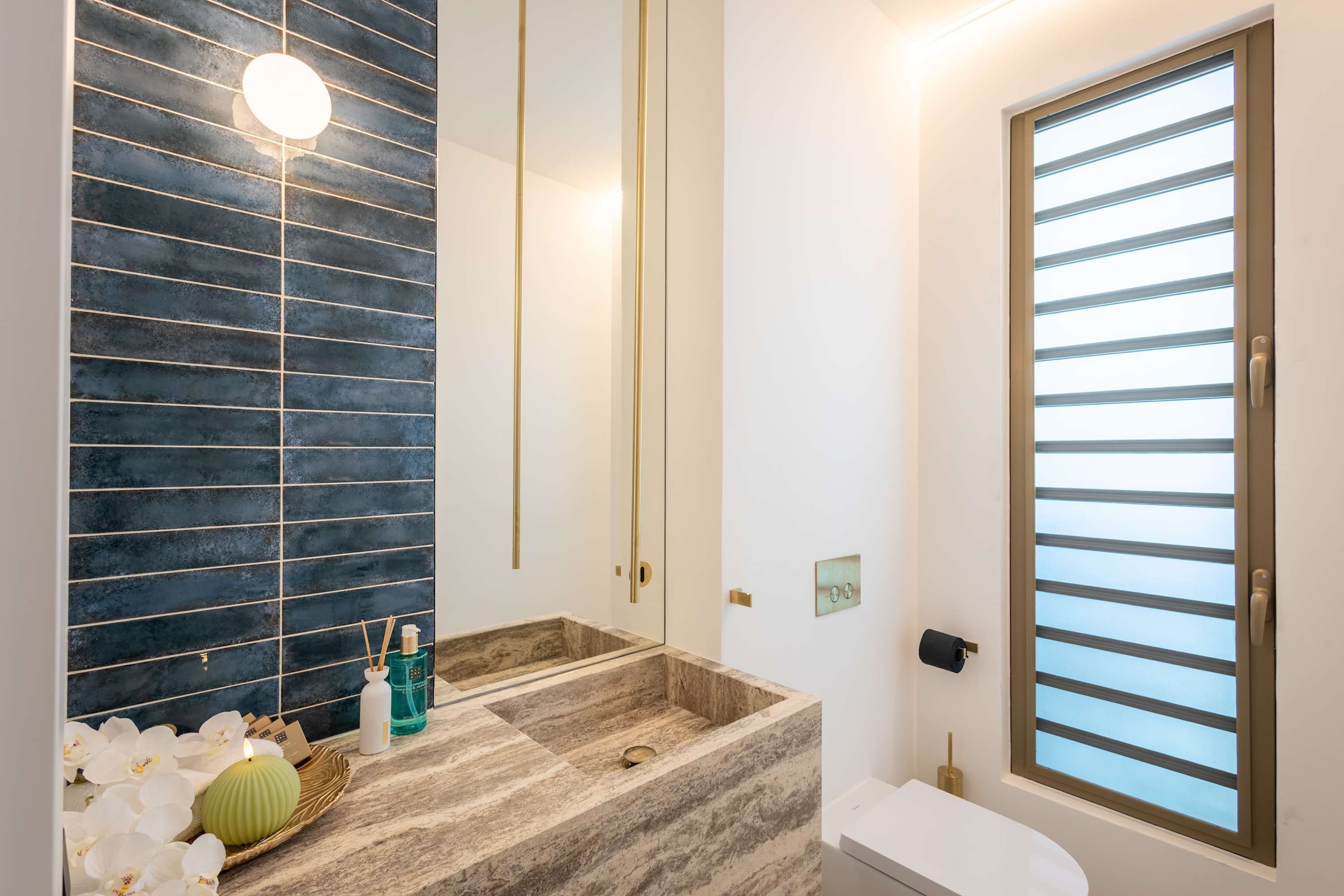 Casa de banho com lavatório de mármore, parede revestida a azulejos escuros, janela com estores e espelho.