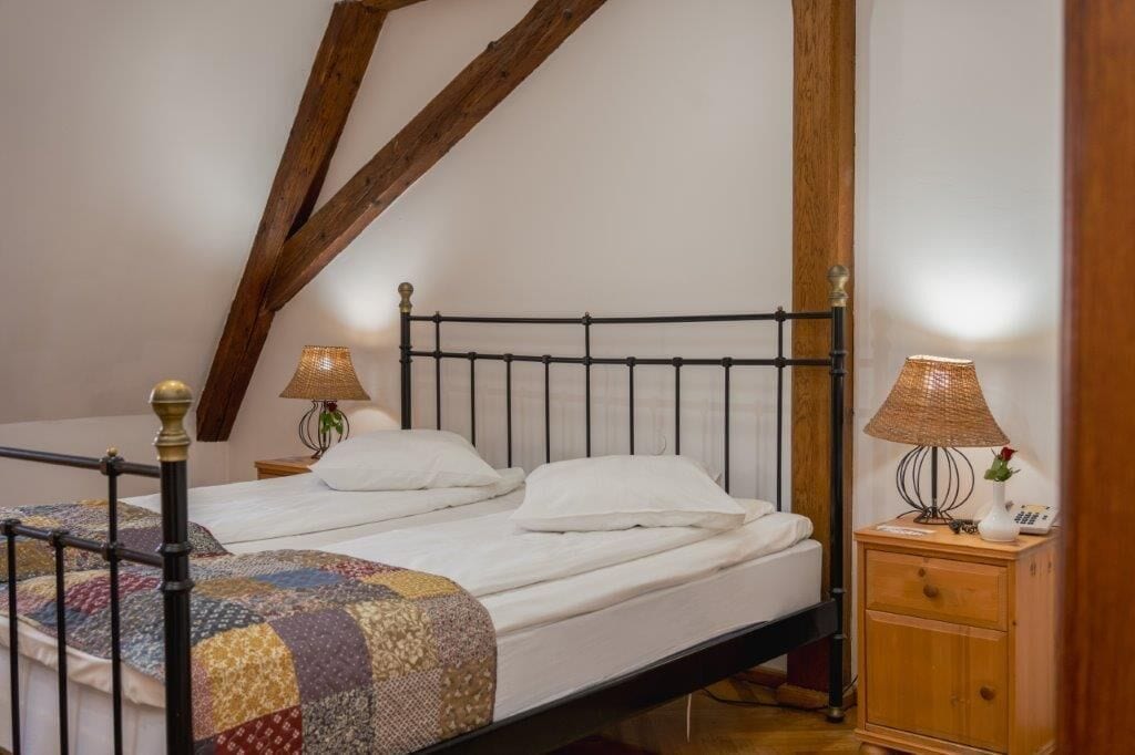 Uma cama com estrutura de metal, roupa de cama branca e um edredom. Há duas luminárias em mesas de cabeceira ao lado da cama.