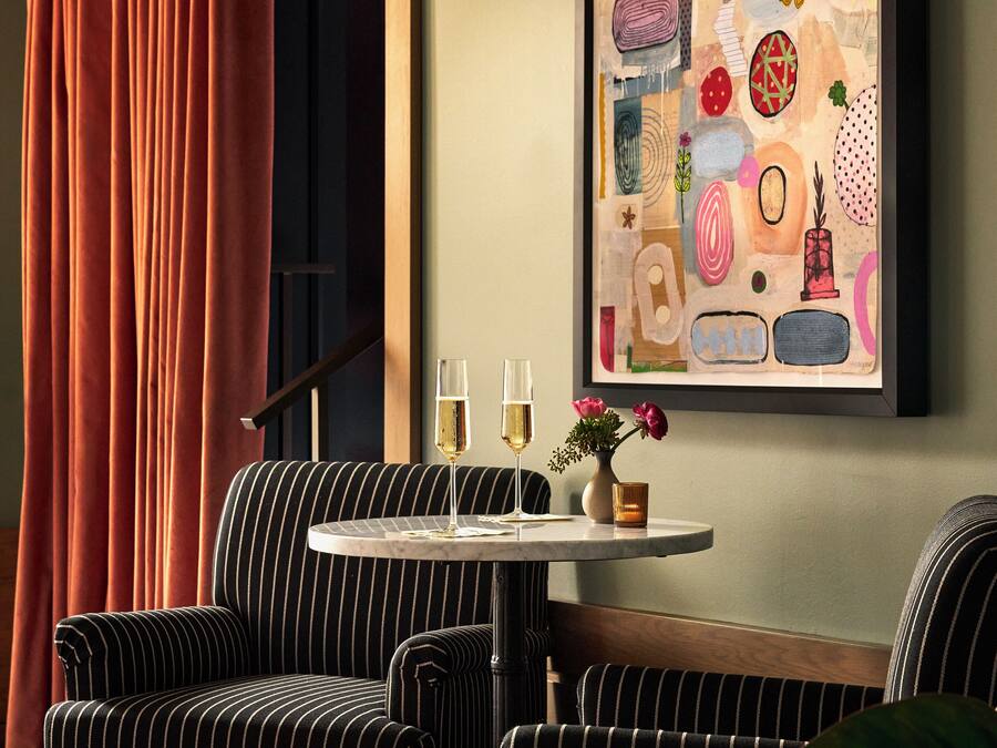 Quarto com cadeiras estofadas listradas, uma pequena mesa redonda com duas taças de vinho branco e um quadro abstrato na parede.