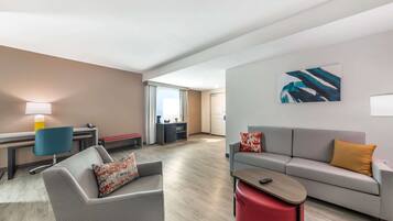Quarto de hotel moderno com sofá cinza, um banquinho vermelho, escrivaninha com luminária e uma grande pintura abstrata na parede.