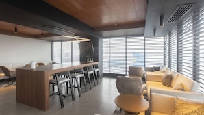 Interior moderno com área de bar central, espaços para sentar e amplas janelas que proporcionam luz natural.