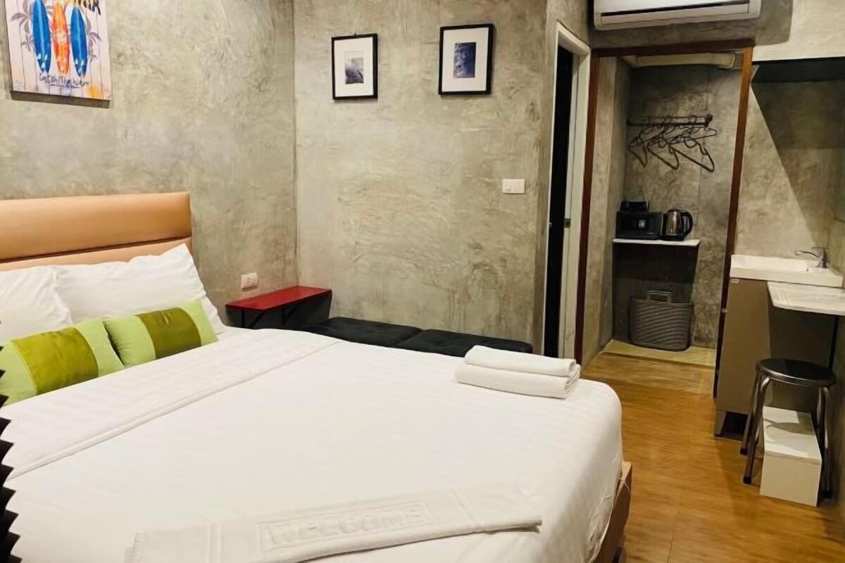 Quarto de hotel com cama, escrivaninha, cadeira, um banquinho pequeno, uma mesa de cabeceira vermelha, uma prateleira na parede com uma chaleira e uma lixeira.