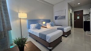 Quarto de hotel com duas camas, mesa de cabeceira, luminária, planta e vista para o mar.