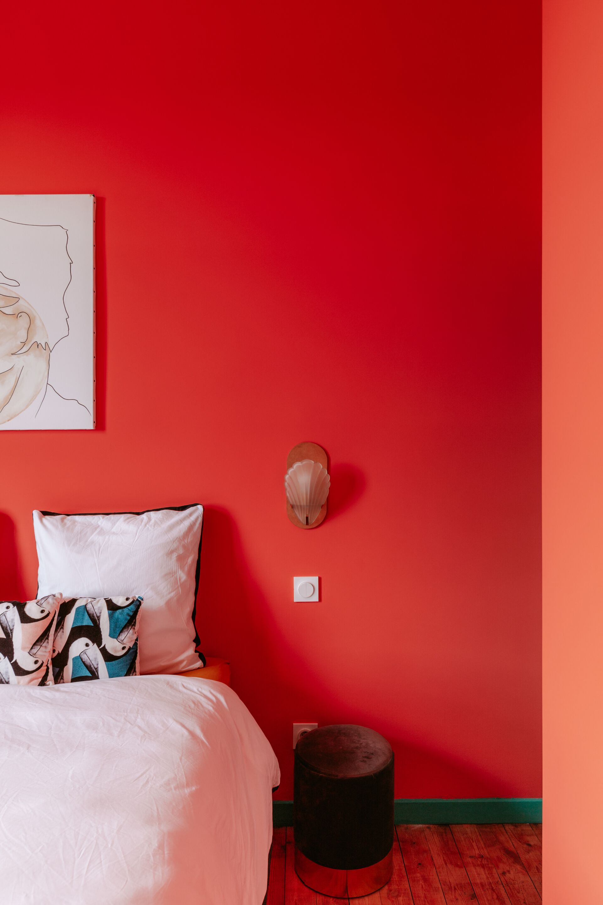 Quarto com parede vermelha, cama com roupa de cama branca, travesseiro branco, um banquinho preto e luminária fixada na parede.