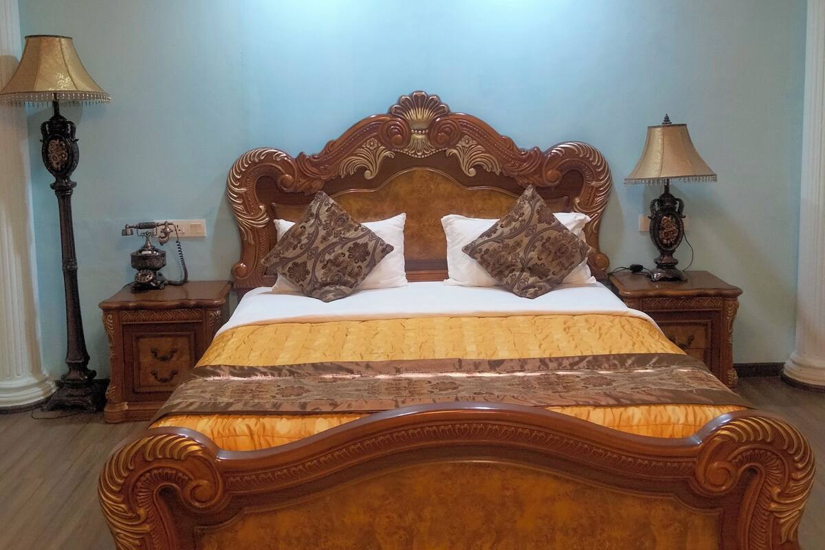 Uma cama grande e ricamente decorada, com estrutura de madeira e colcha amarela. Há duas mesas de cabeceira com luminárias de cada lado da cama. O quarto tem paredes azul-claras e piso de madeira.