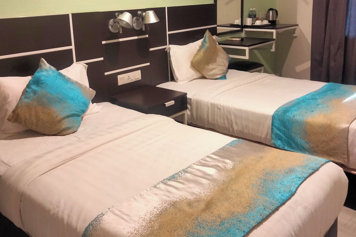 Quarto de hotel com duas camas, cada uma com colcha branca e manta azul e bege. Apresenta uma mesa de cabeceira com luminária, espelho e uma pequena mesa com chaleira e chávenas.