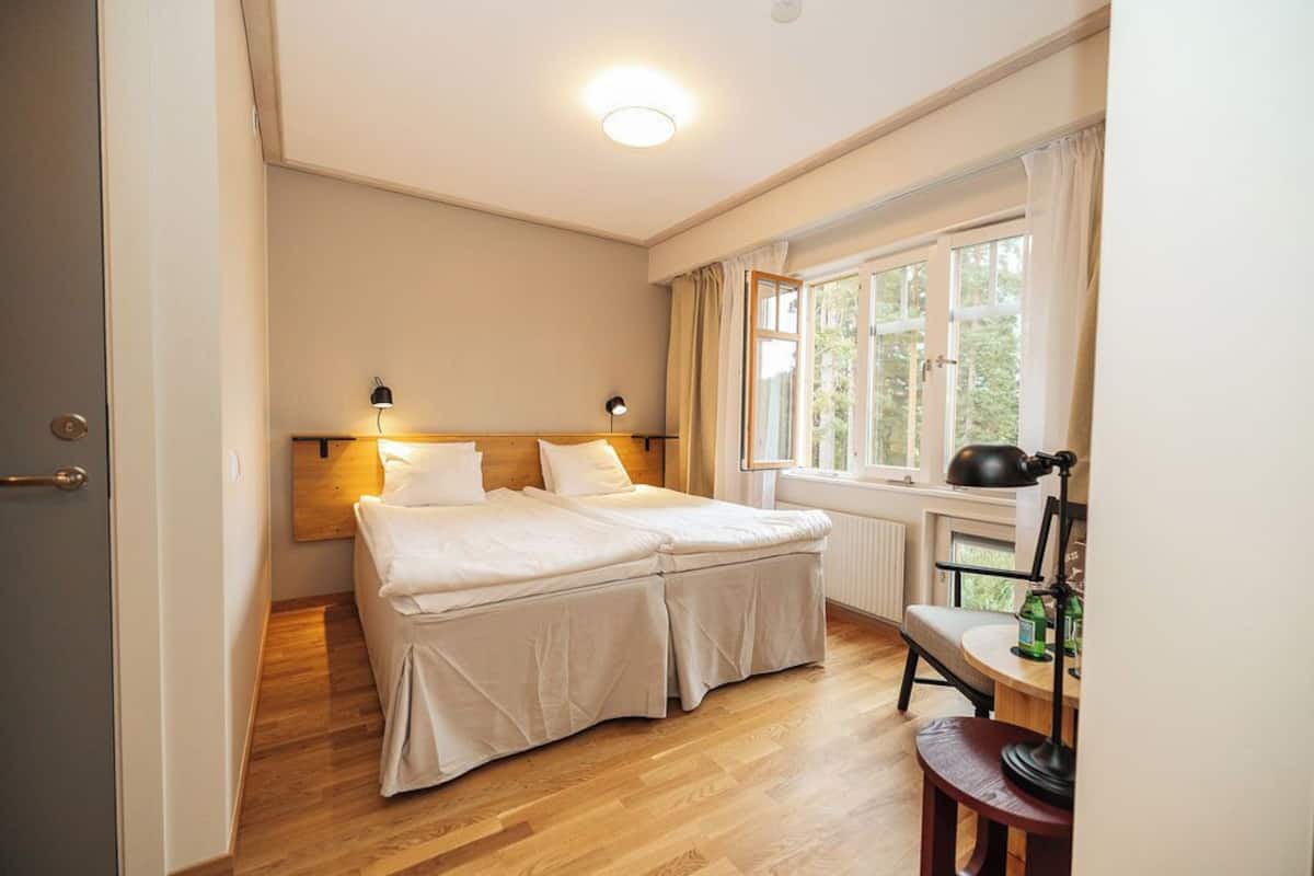 Quarto com duas camas, cabeceira de madeira, luminárias de cabeceira, janela com cortinas, piso de madeira e uma pequena mesa com cadeira.